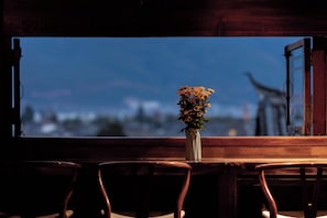 Uma mesa de madeira com um vaso de flores em frente a uma janela com uma persiana aberta.
