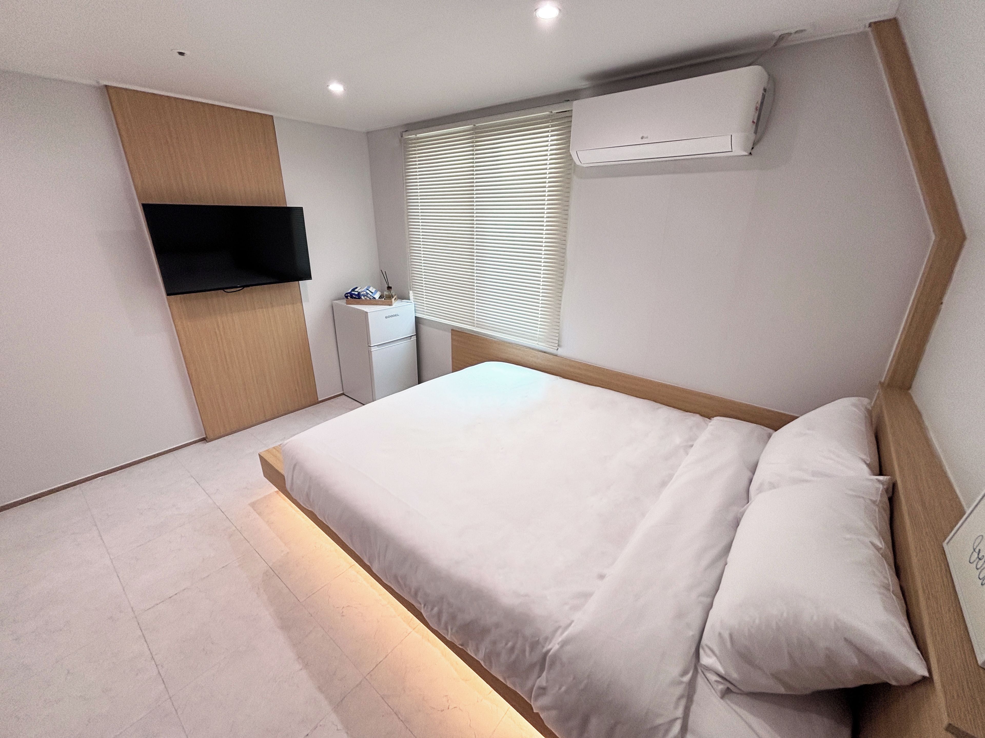 Quarto com cama, televisão, pequeno espaço de cozinha e janela com persianas.