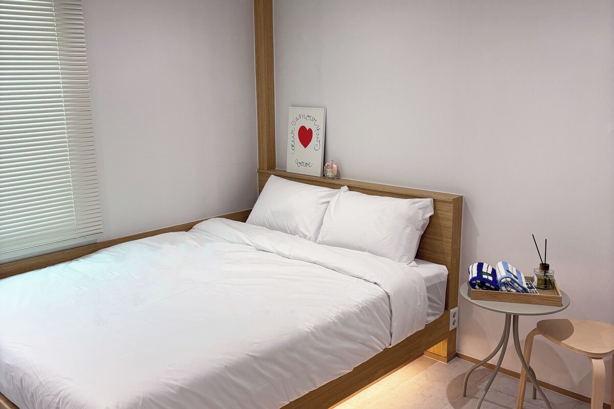Uma cama bem arrumada com lençóis brancos, uma mesa de cabeceira com abajur e um quadro fixado na parede.