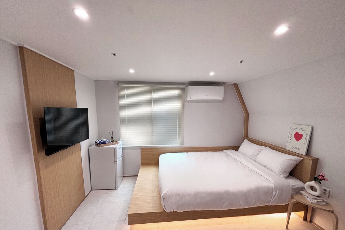 Quarto com cama, mesa de cabeceira, televisão e janela com persianas.