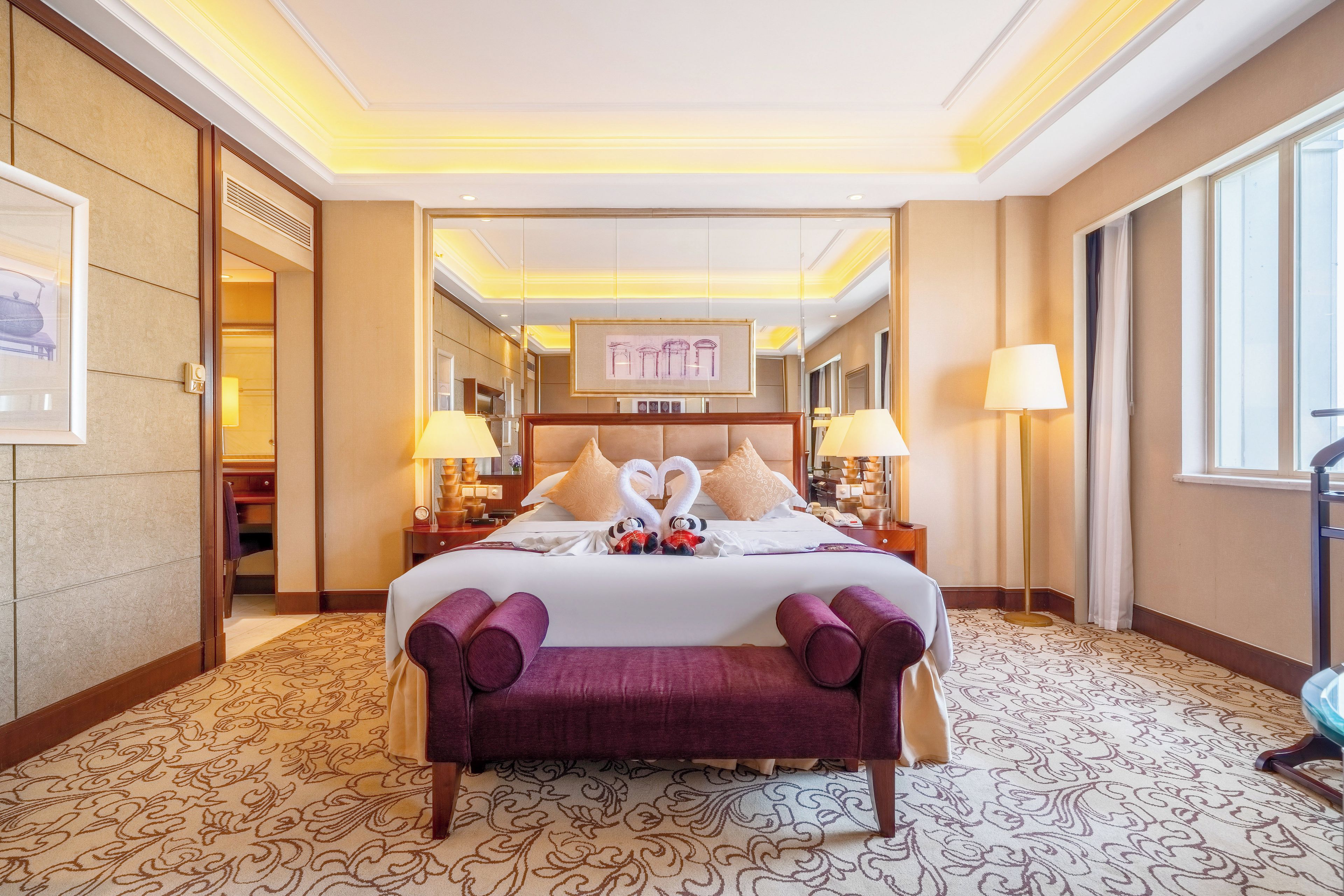 Quarto de hotel com uma cama grande, um banquinho, uma escrivaninha e um espelho.