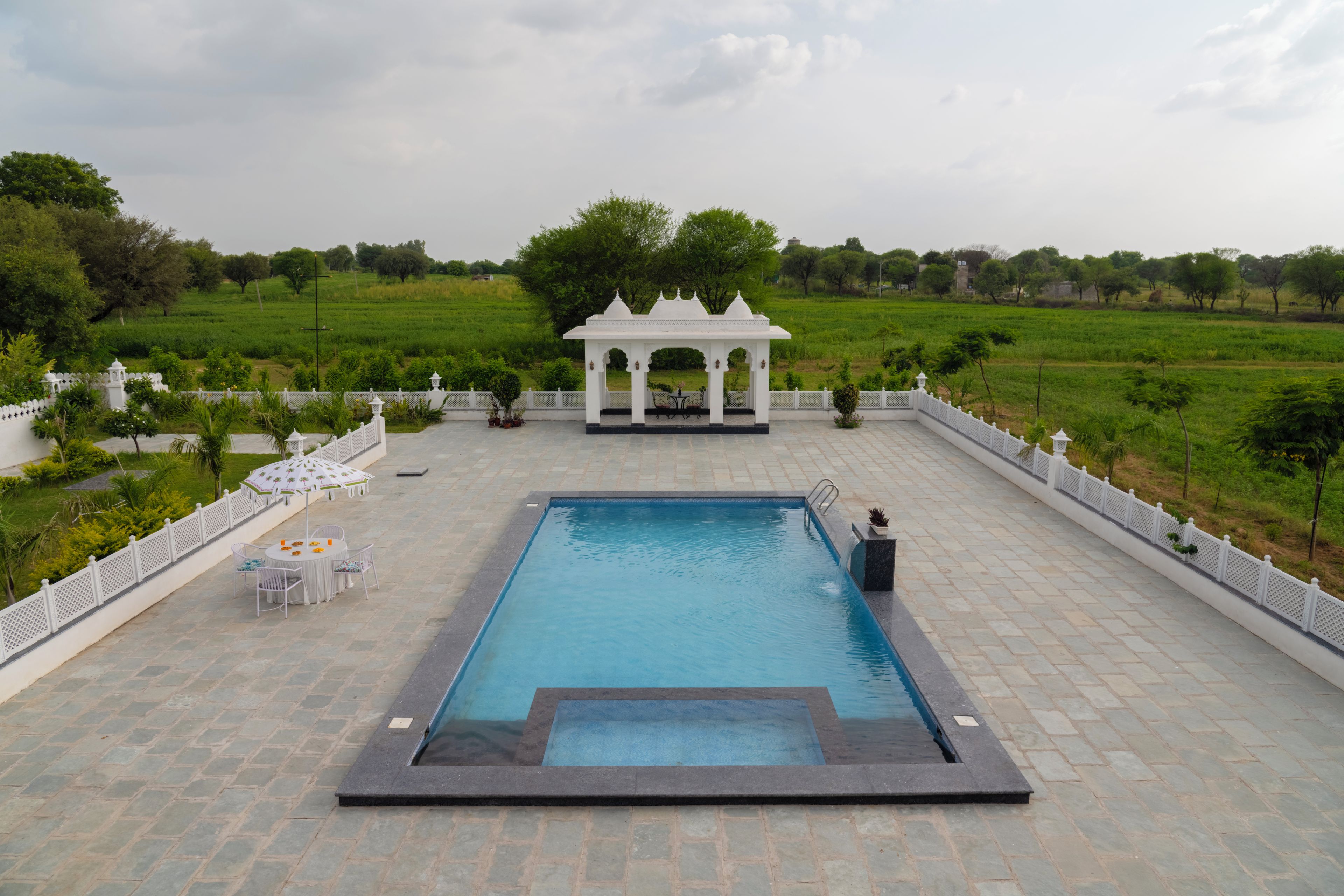 Piscina retangular com um gazebo branco ao fundo, rodeada por uma cerca branca e vegetação.