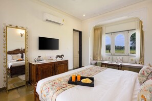 Quarto de hotel com cama, televisão, escrivaninha, janela com cortinas e espelho com moldura decorativa.