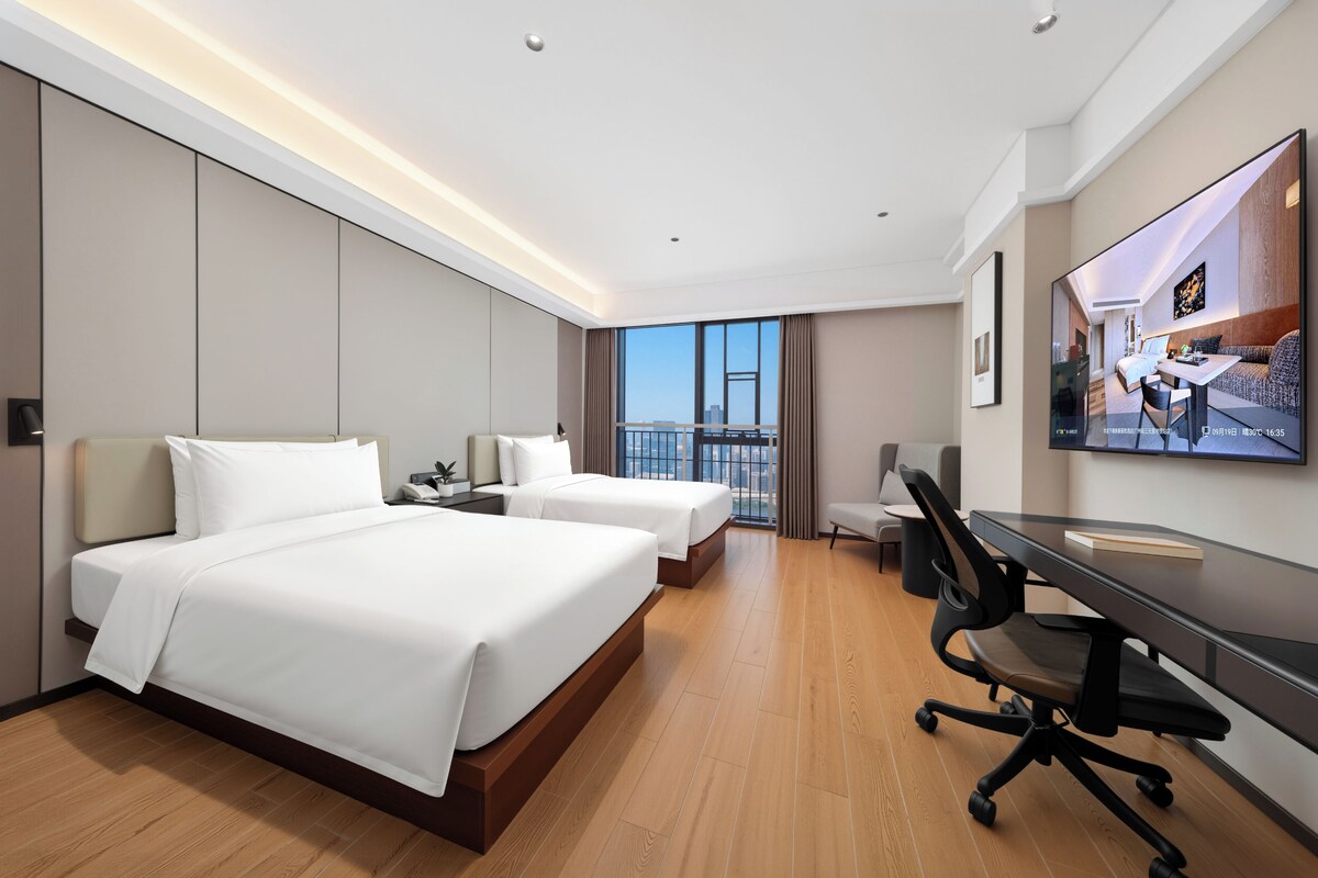 Quarto de hotel com duas camas, uma escrivaninha, uma cadeira, televisão e varanda com vista para a cidade.