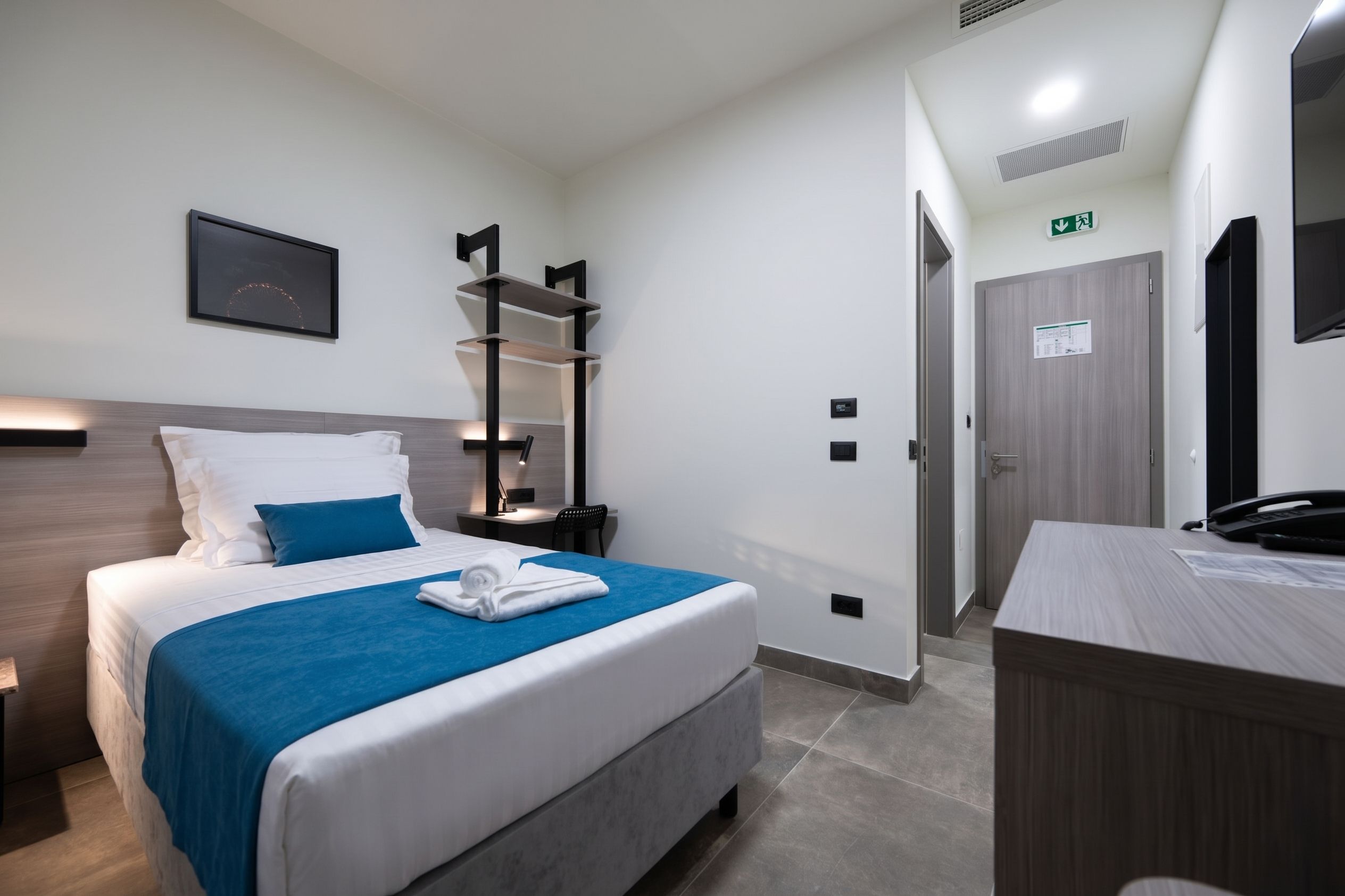Quarto de hotel com cama, escrivaninha, cadeira, televisão e porta do banheiro.