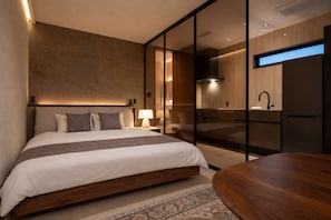 Quarto de hotel moderno com uma cama grande, mesa de cabeceira de madeira, escrivaninha embutida e divisória de vidro separando o quarto do banheiro.