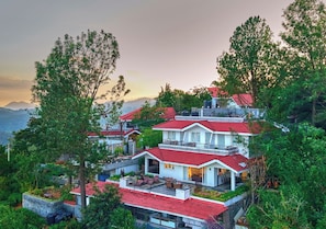 Edifício residencial de vários andares com telhado de telhas vermelhas, cercado por vegetação exuberante e com vista para uma paisagem montanhosa.