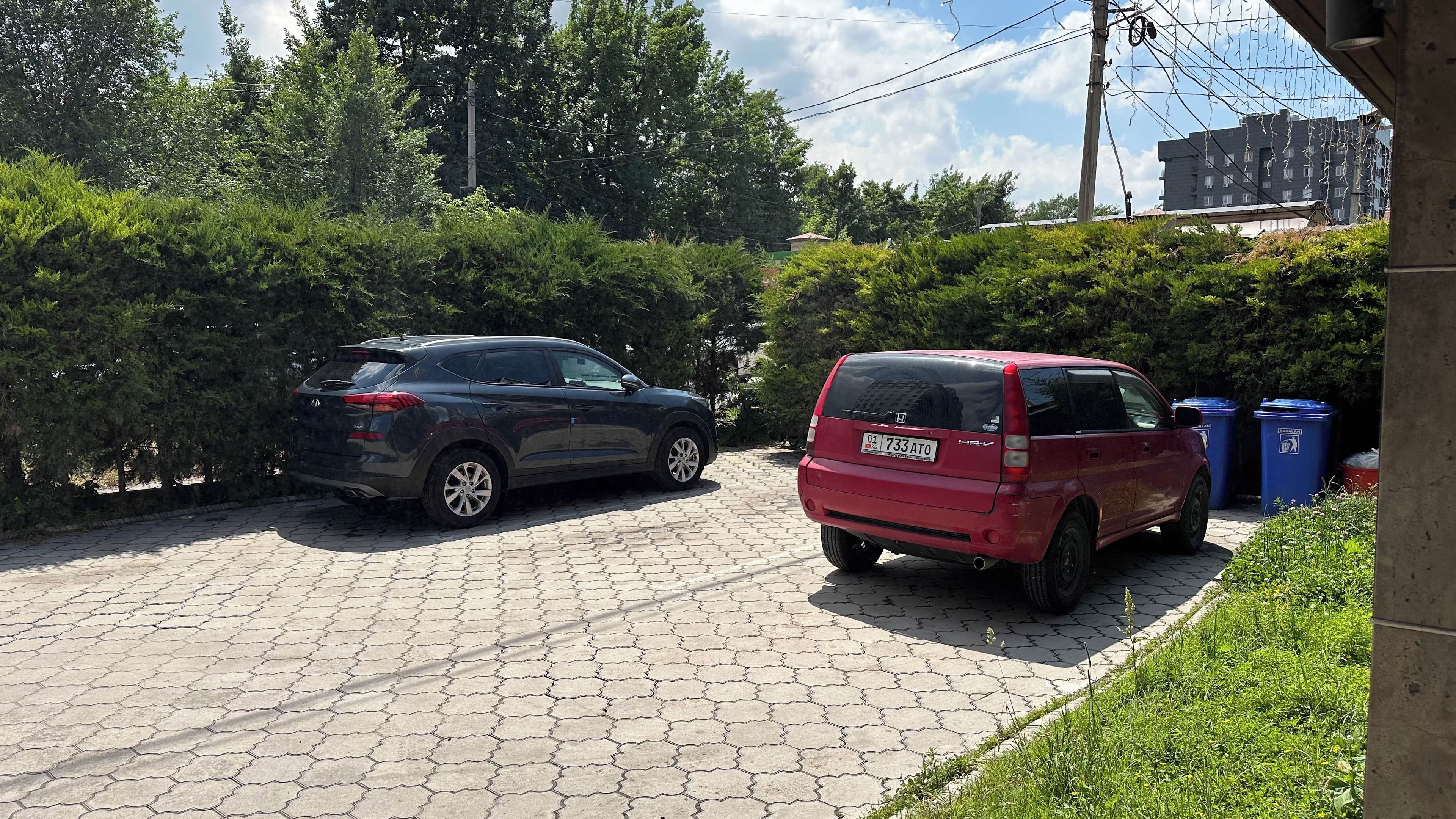 Dois carros estacionados em um caminho de paralelepípedos, com um hedge e árvores ao fundo.
