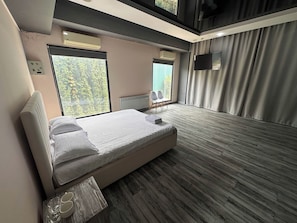 Quarto de hotel moderno com uma cama grande, janela com vista para árvores, radiador e televisão de tela plana fixada na parede.