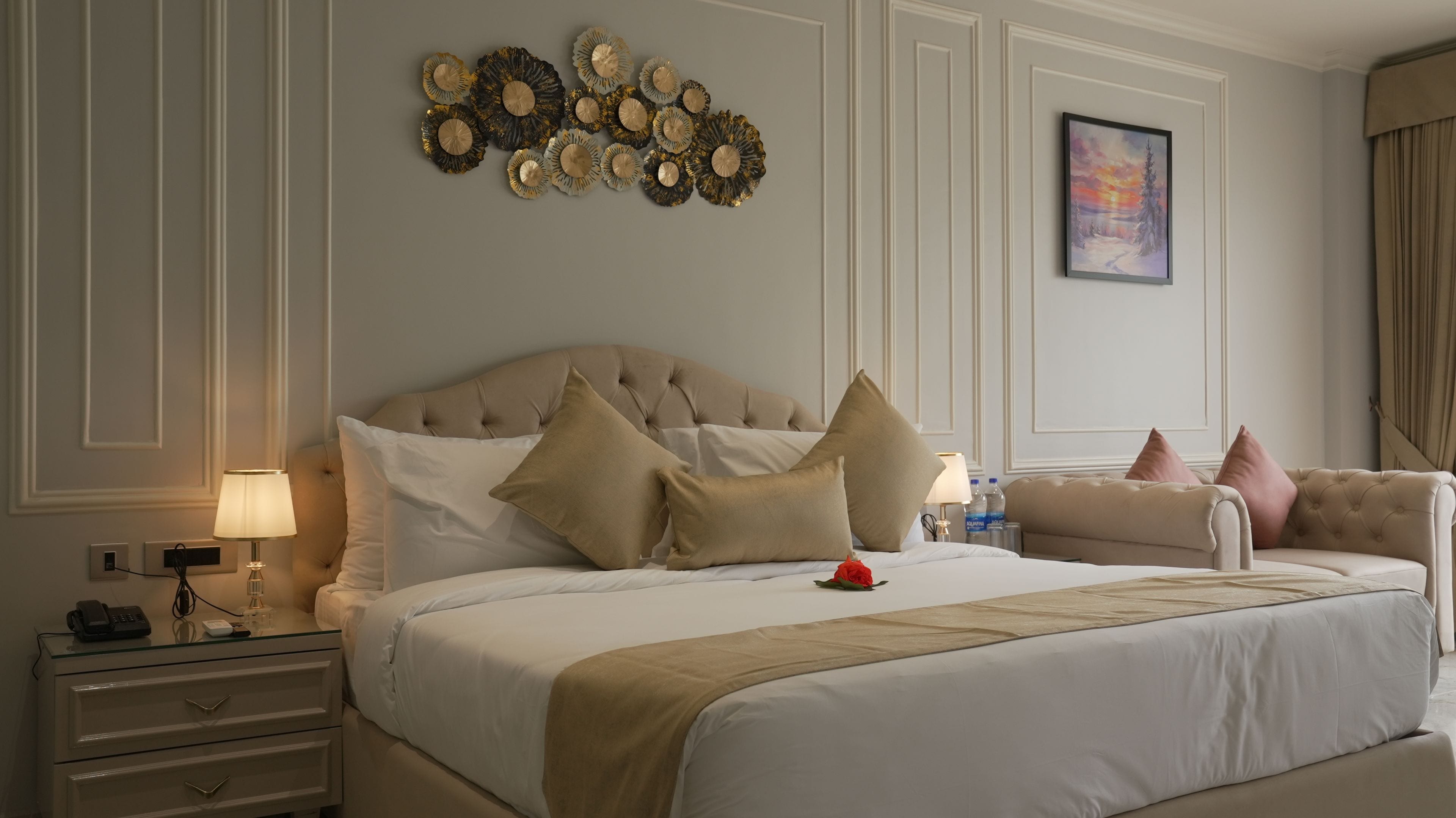 Quarto de hotel com uma cama grande, mesa de cabeceira com abajur, criado-mudo e um quadro na parede.