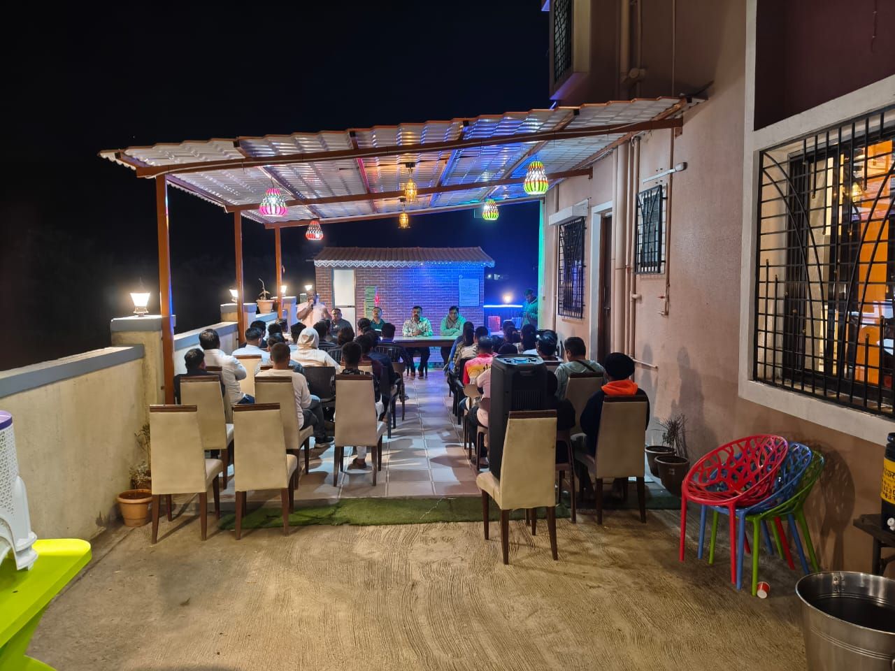 Uma área externa com mesas e cadeiras, um espaço coberto com palco, e um edifício com janelas protegidas por grades.