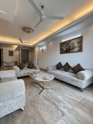 Uma sala de estar moderna com piso de mármore, dois sofás, uma mesa de centro, ventiladores de teto, iluminação embutida e um quadro na parede.