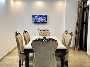 Uma sala de jantar com uma mesa retangular e seis cadeiras, uma TV de tela plana na parede e uma porta com cortinas.