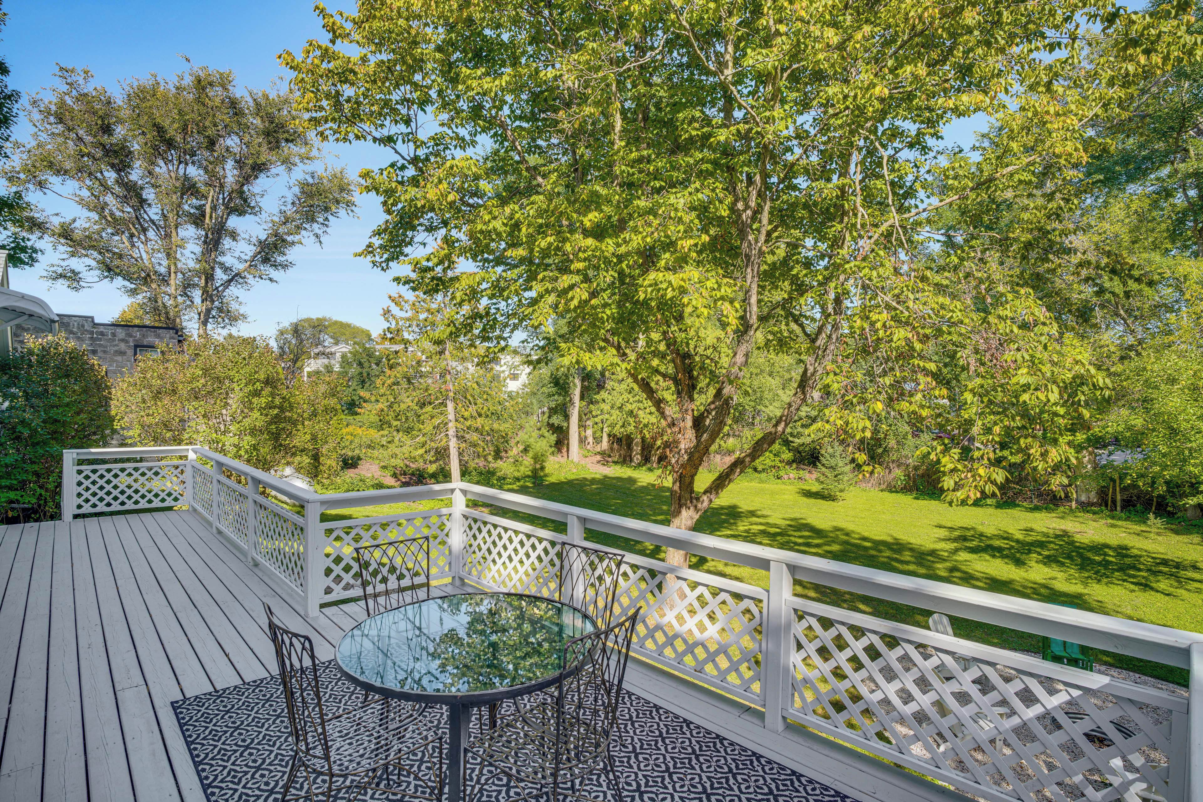 Um terraço com mesa e cadeiras de vidro, cercado por um guarda-corpo de treliça branca, com vista para um gramado bem cuidado e árvores.