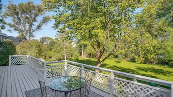 Um terraço com mesa e cadeiras de vidro, cercado por um guarda-corpo de treliça branca, com vista para um gramado bem cuidado e árvores.