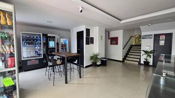 Um hall limpo com máquinas de venda automática, uma mesa com cadeiras, uma lixeira e uma escada.