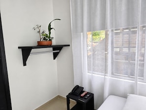 Quarto de hotel com cama, mesa de cabeceira pequena com telefone e controles remotos, janela com cortinas leves e prateleira na parede com planta em vaso.