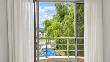 Vista pela janela, com cortinas brancas, mostrando uma varanda com guarda-sol azul e vegetação.