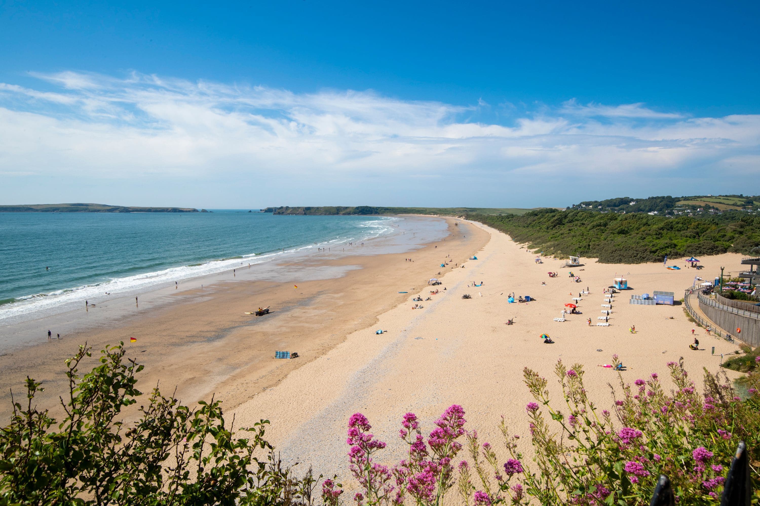 Uma extensa praia de areia com céu claro, mar calmo e paisagem montanhosa ao longe.