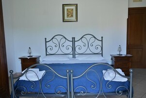 Uma cama com estrutura de metal, colcha azul e lençóis brancos. Há duas mesas de cabeceira com abajures.