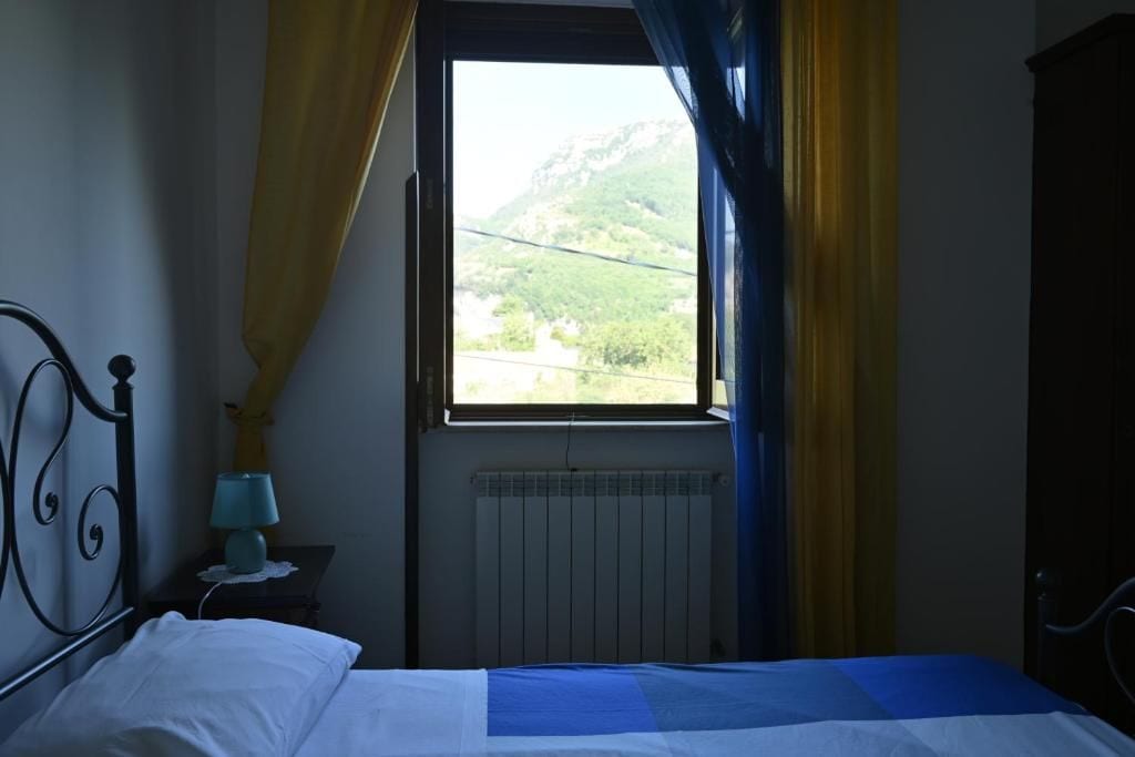 Quarto com cama, janela com cortinas, radiador e mesa de cabeceira com abajur.