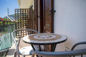 Uma pequena mesa de exterior com tampo de azulejos, rodeada por cadeiras de metal, encostada a uma parede com portadas de madeira.