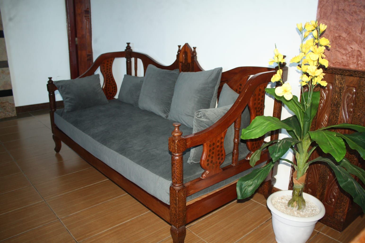 Um sofá-cama de madeira com almofadas cinzentas, uma planta em vaso com flores amarelas e um chão revestido de azulejos.