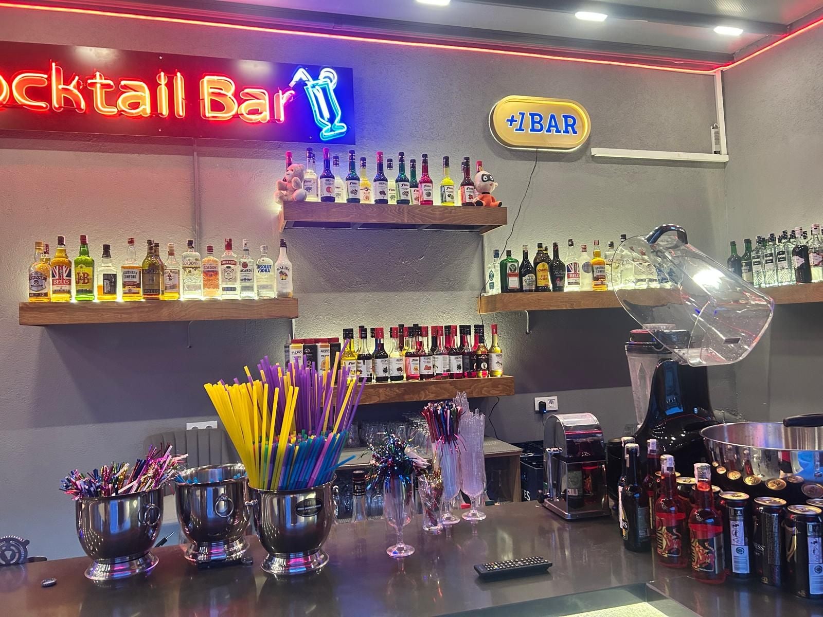 Uma área de bar com prateleiras de garrafas de bebidas, um letreiro de neon e uma máquina de café.