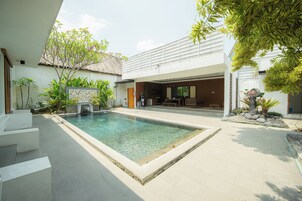 Uma área de piscina com superfície de água cristalina, cercada por um edifício branco com telhado de palha e uma área de estar coberta ao ar livre.