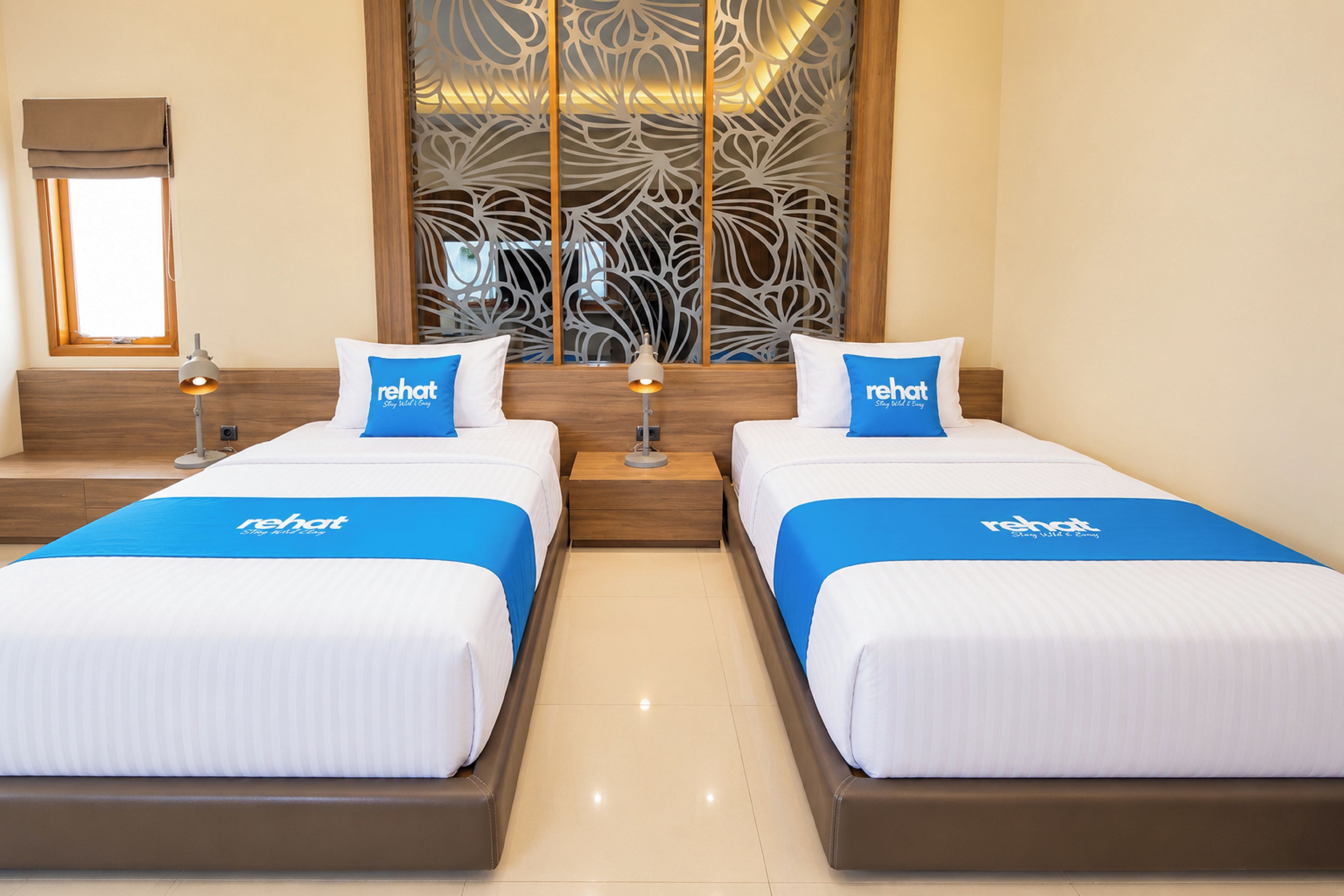 Quarto de hotel com duas camas, cada uma com colcha azul e almofadas brancas, e cabeceira de madeira decorativa.