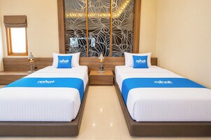 Quarto de hotel com duas camas, cada uma com colcha azul e almofadas brancas, e cabeceira de madeira decorativa.