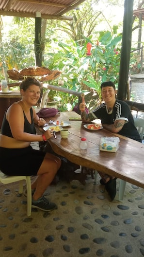 Duas pessoas sentadas à mesa de madeira com comida e bebida, rodeadas por vegetação.