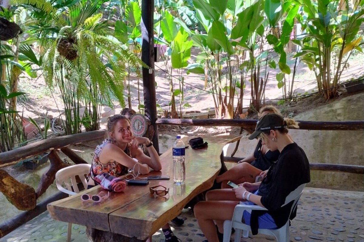 Uma área de estar coberta ao ar livre com uma mesa rústica de madeira, rodeada por vegetação exuberante.