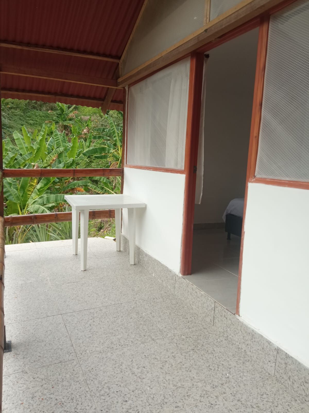 Varanda coberta com mesa e cadeiras brancas, vista para o verde e porta que dá para um espaço interno.