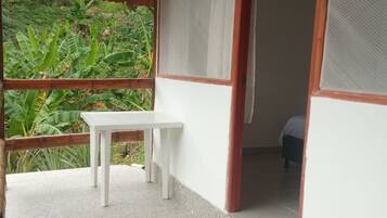 Varanda coberta com mesa e cadeiras brancas, vista para o verde e porta que dá para um espaço interno.