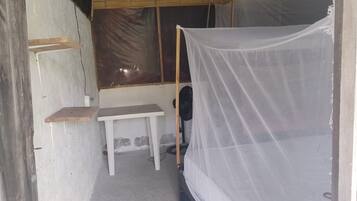 Pequeno quarto privativo com uma cama de solteiro, um banco e uma mesa.