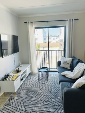 Uma sala de estar moderna com televisão de tela plana, um tabuleiro de xadrez, um sofá com almofadas, uma mesinha lateral e um tapete estampado.