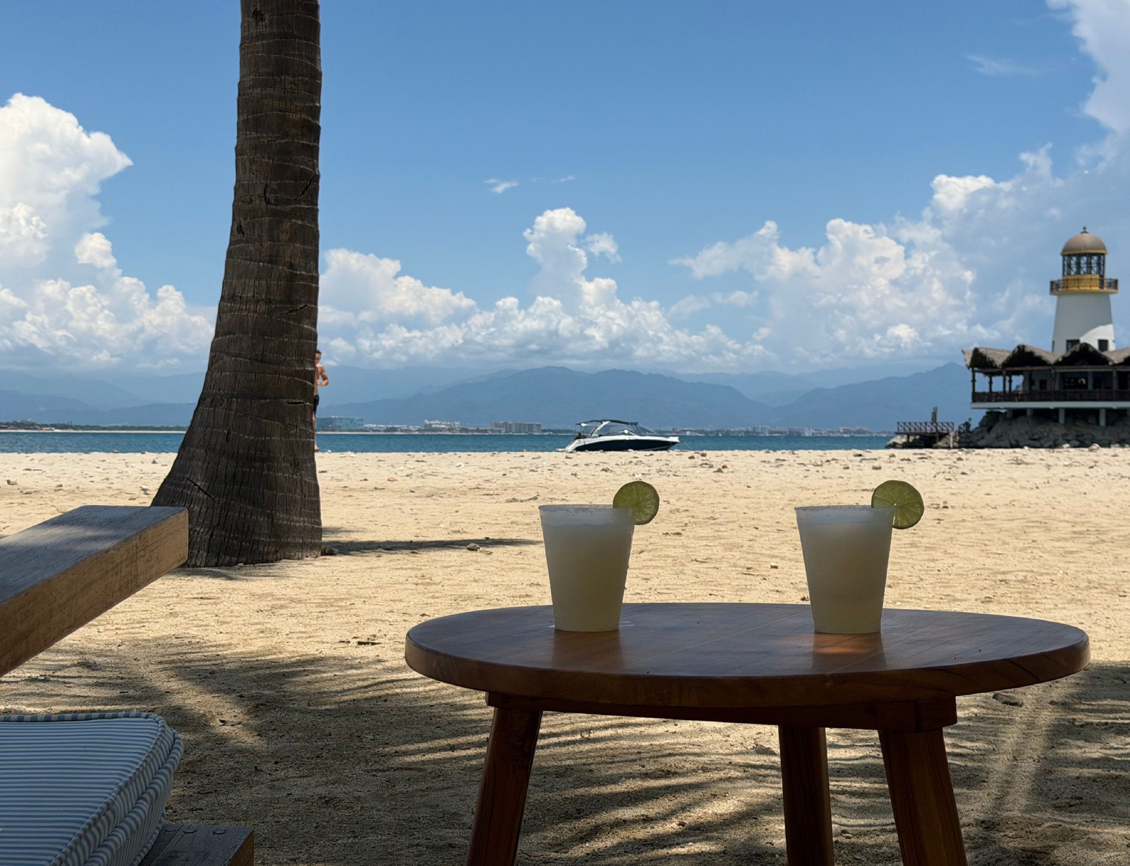 Uma mesa à beira-mar com duas bebidas, uma palmeira e um farol ao fundo.