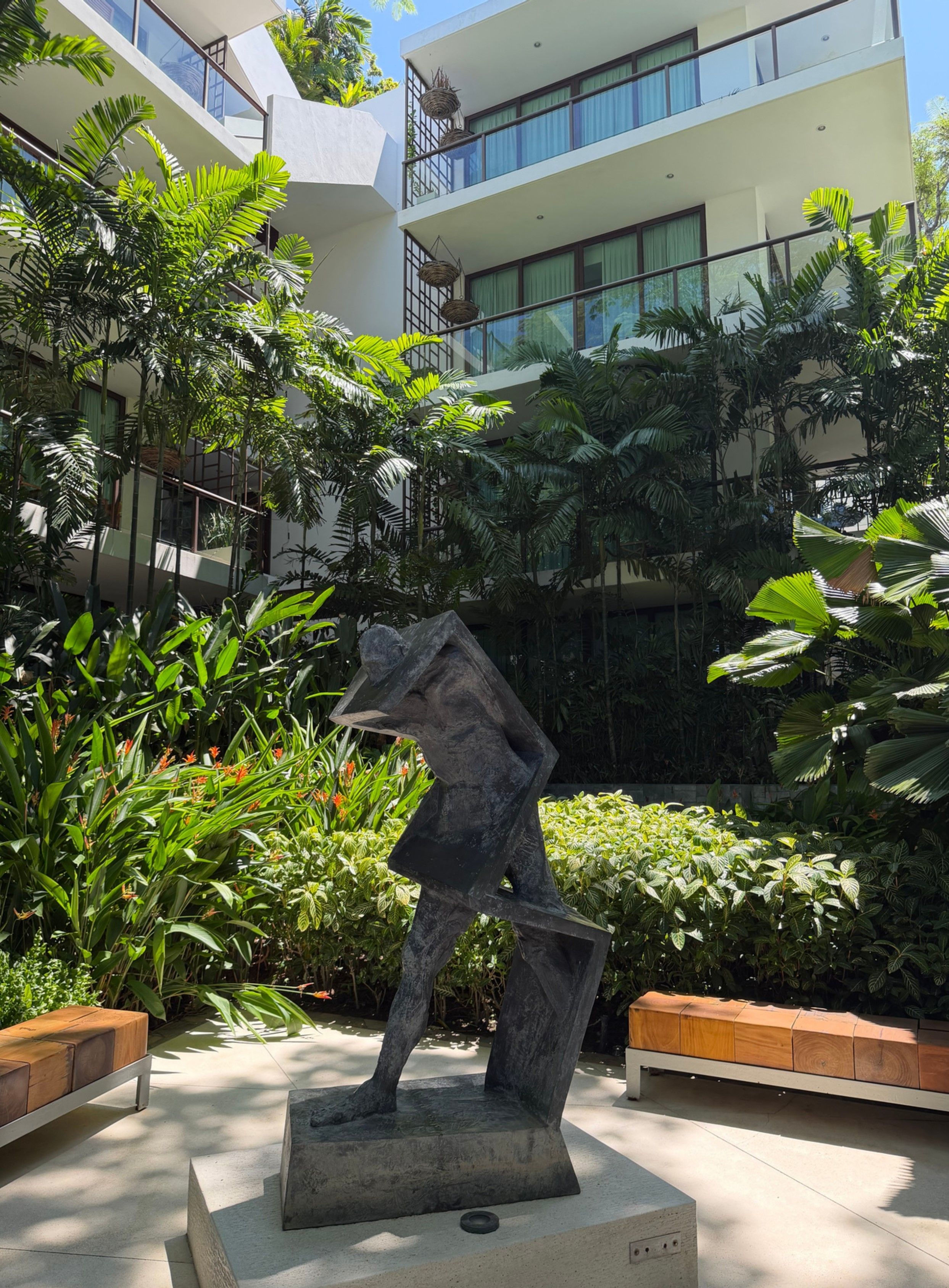 Escultura em um pátio cercado por vegetação exuberante e um edifício moderno com sacadas.