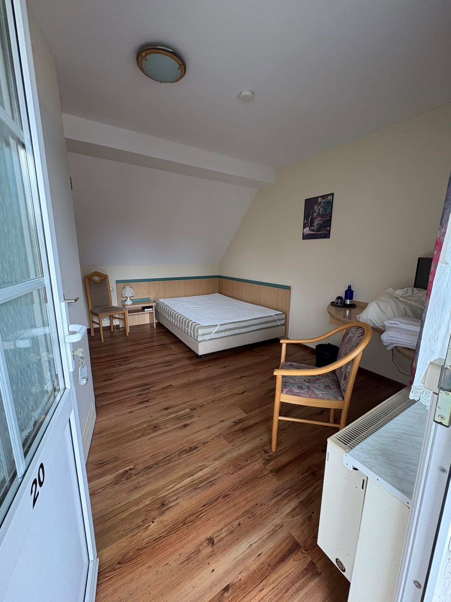 Acomodação pequena, com um quarto único, piso de madeira, cama, escrivaninha com cadeira, radiador e janela.