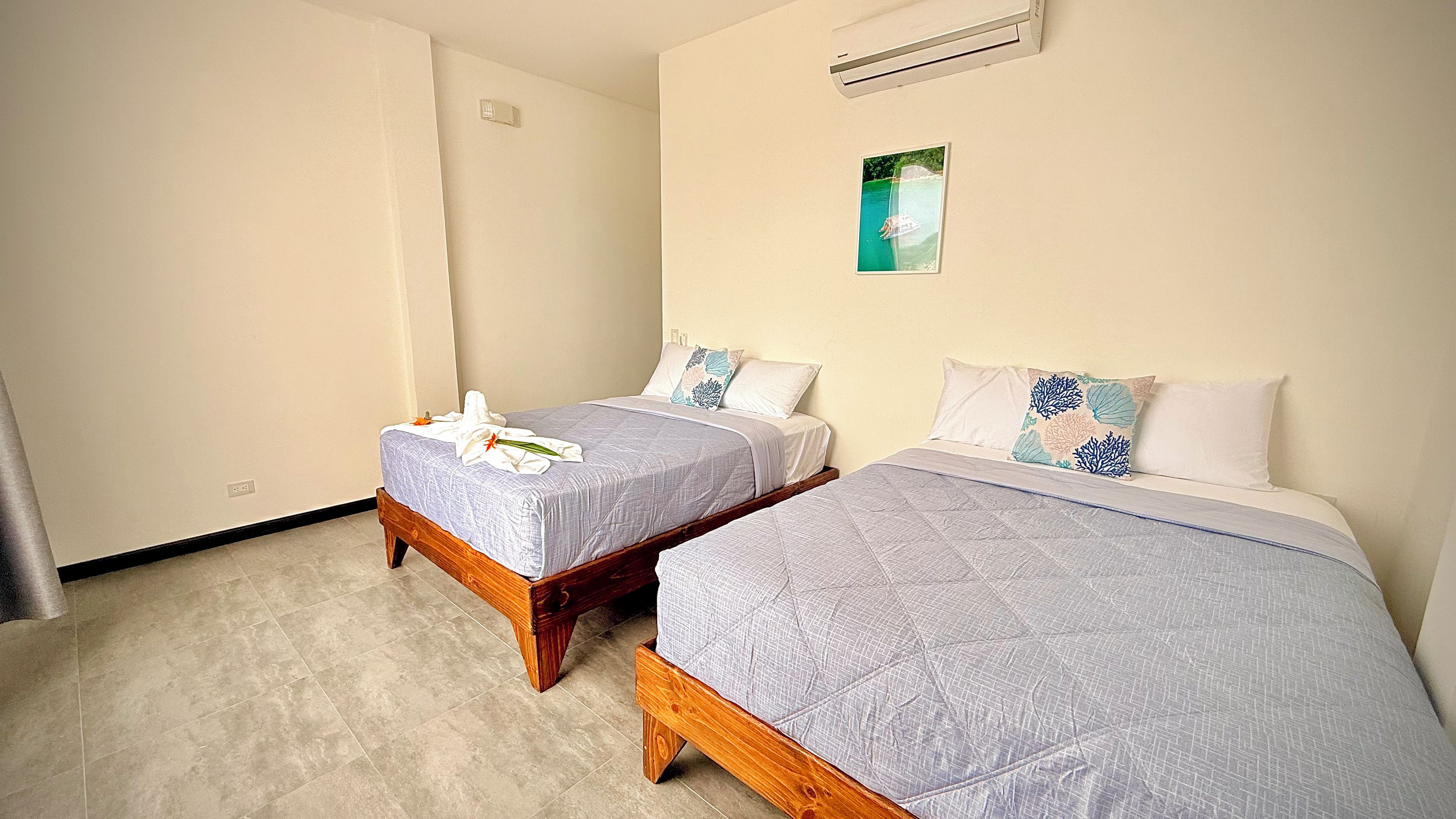 Quarto de hotel com duas camas, cada uma com estrutura de madeira e roupa de cama cinzenta. Há uma pequena imagem na parede acima das camas.