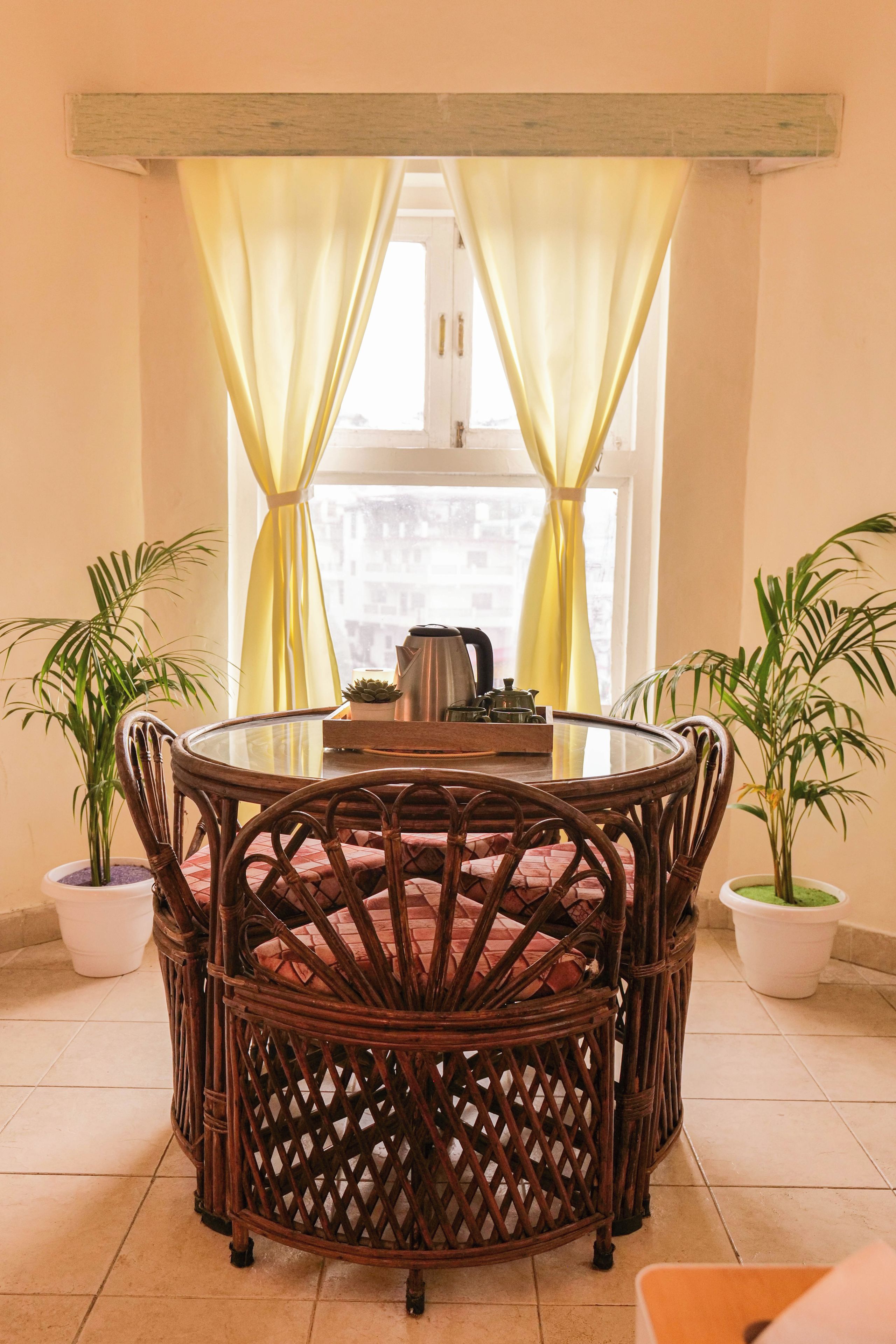 Uma mesa redonda de vime com tampo de vidro, acompanhada por duas plantas em vasos, num quarto com janela e cortinas amarelas.