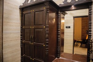 Uma porta dupla de madeira com puxadores de latão e detalhes decorativos, dando acesso a um espaço com parede de mármore e outra porta com uma placa em cima.