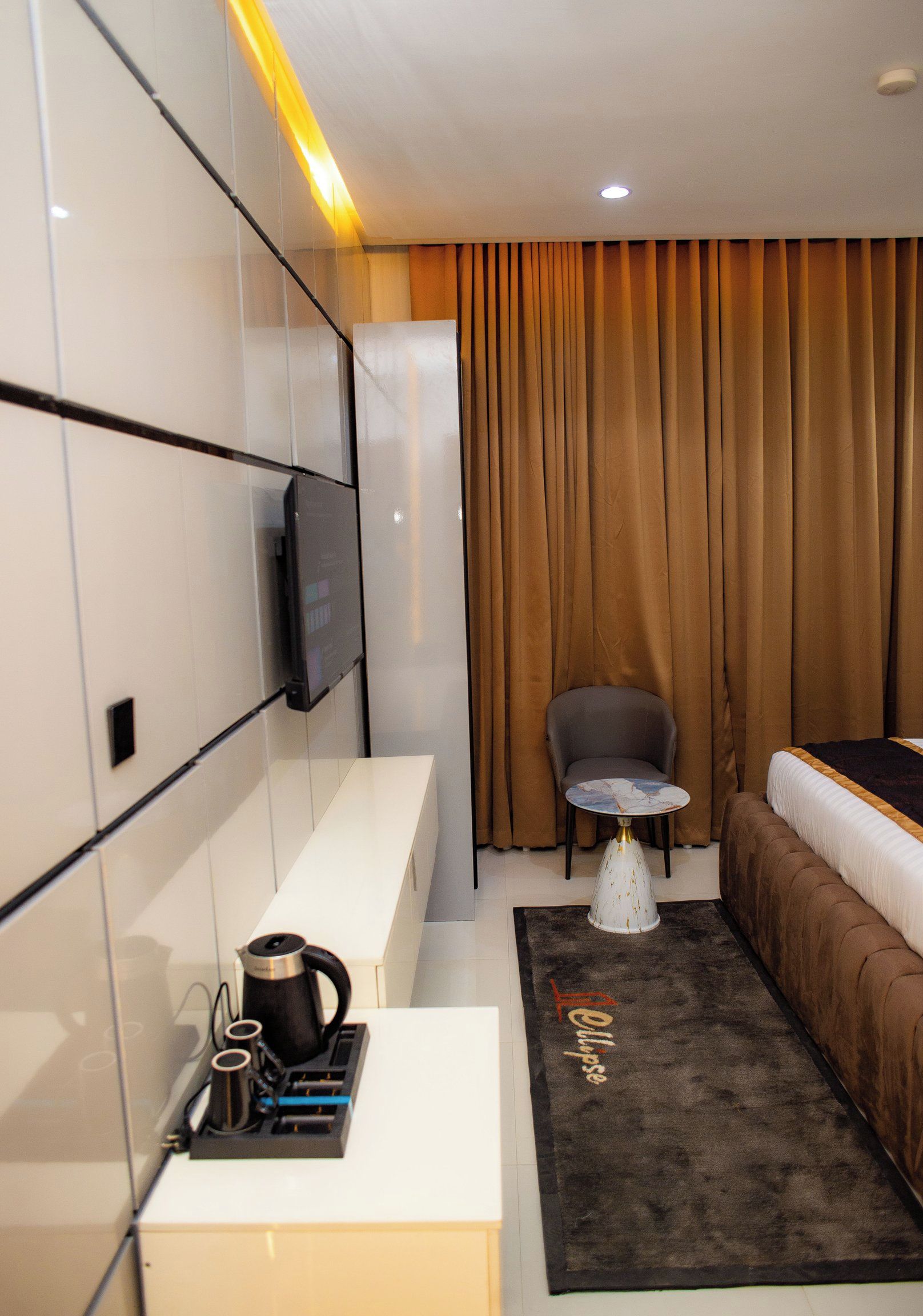 Quarto de hotel com televisão de tela plana na parede, um sofá, uma pequena mesa redonda, uma cafeteira e um tapete com a palavra 'Eclipse'.