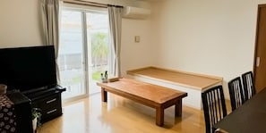 Uma sala de estar com uma mesa de madeira, televisão e uma porta de vidro corria para uma varanda.