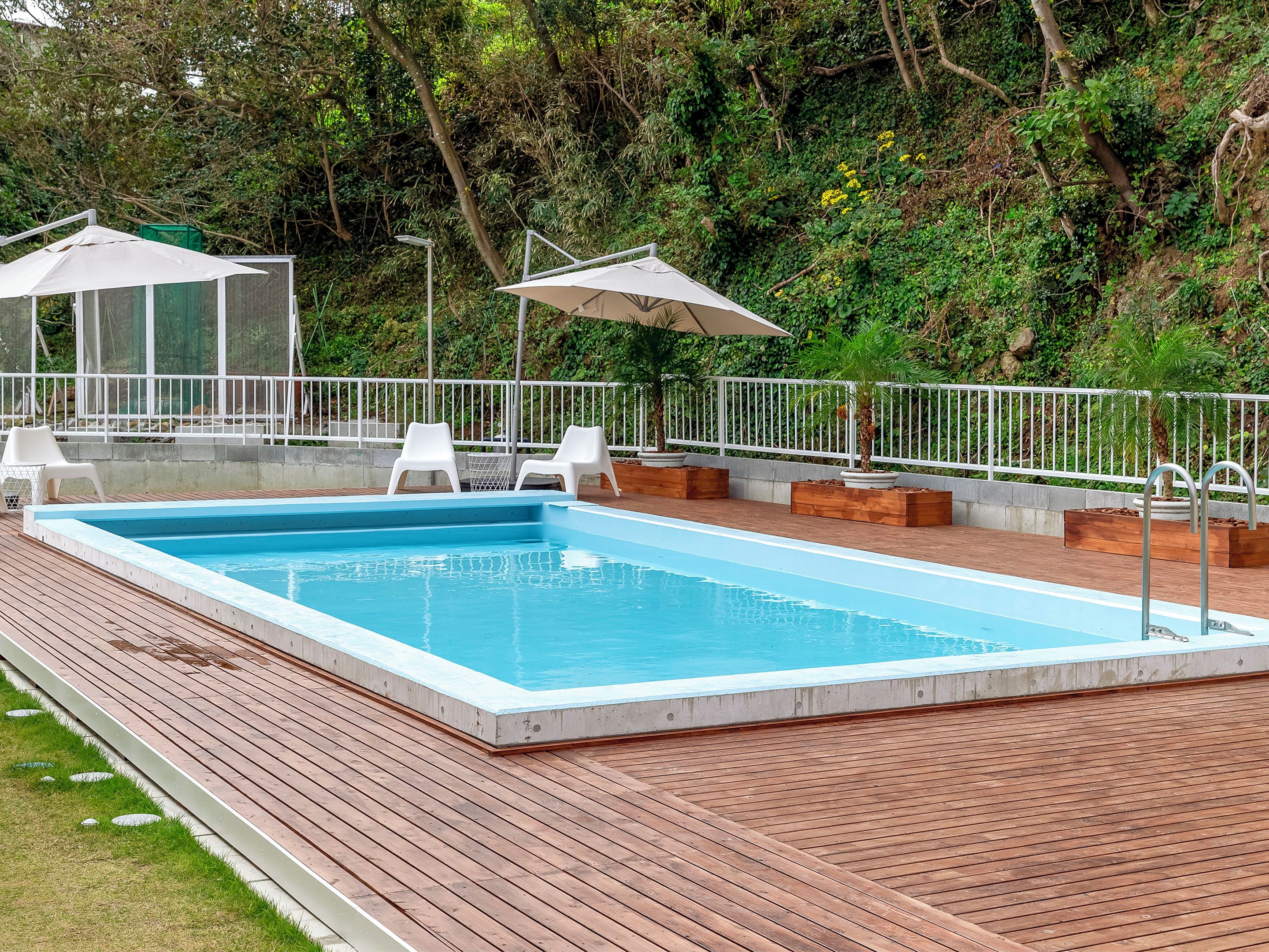 Piscina retangular com deck de madeira, rodeada por cadeiras e guarda-sóis brancos, com um fundo de vegetação densa.