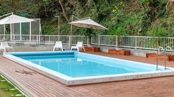 Piscina retangular com deck de madeira, rodeada por cadeiras e guarda-sóis brancos, com um fundo de vegetação densa.