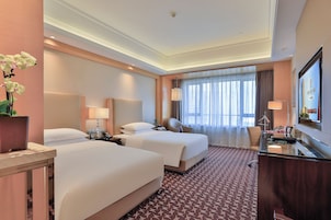 Quarto de hotel com duas camas, uma mesa de trabalho com telefone, espelho, televisão e janela com cortinas.