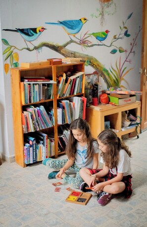 Duas crianças brincam com brinquedos em um chão de azulejos, cercadas por estantes cheias de livros e um mural com pássaros na parede.