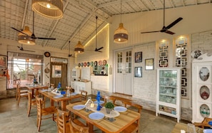 Um restaurante com mesas e cadeiras de madeira, lustres de vime pendurados e uma parede de tijolos brancos com quadros emoldurados.